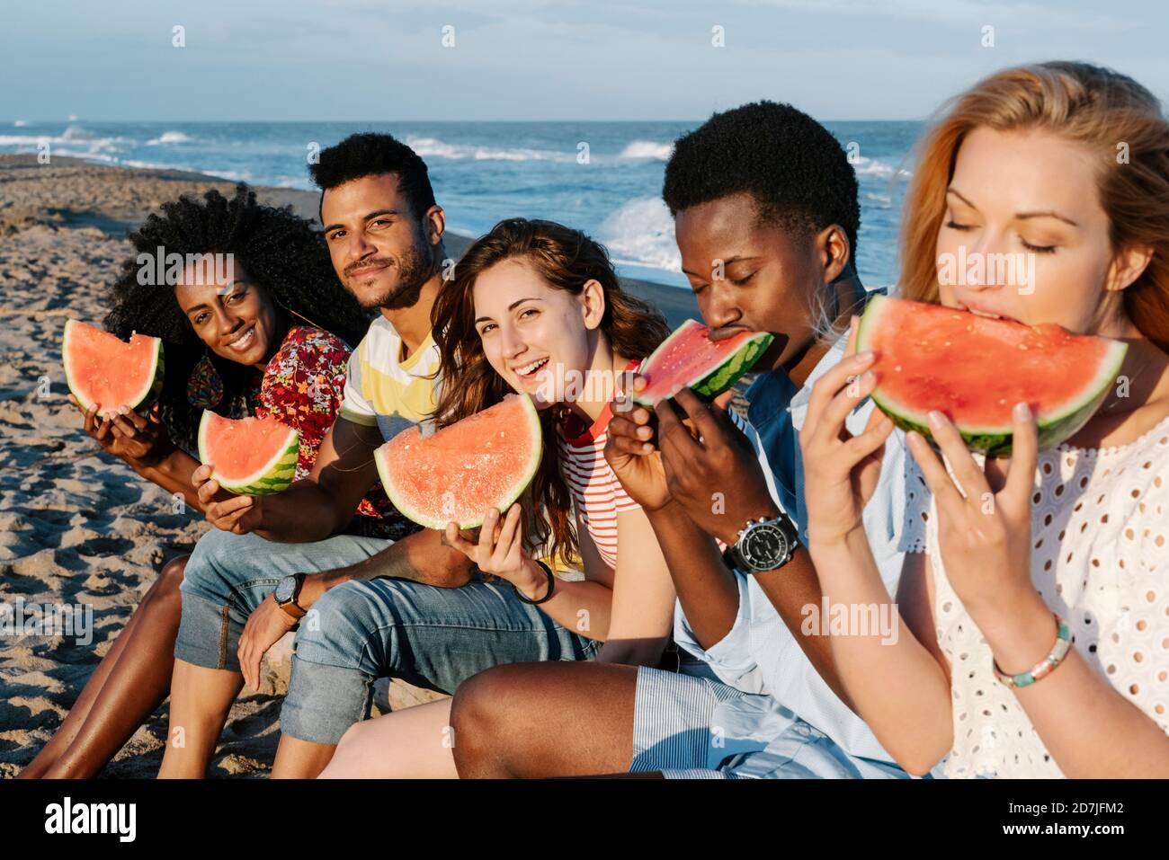 Friends eating slice of watermelon while sitting on beach during sunny ...