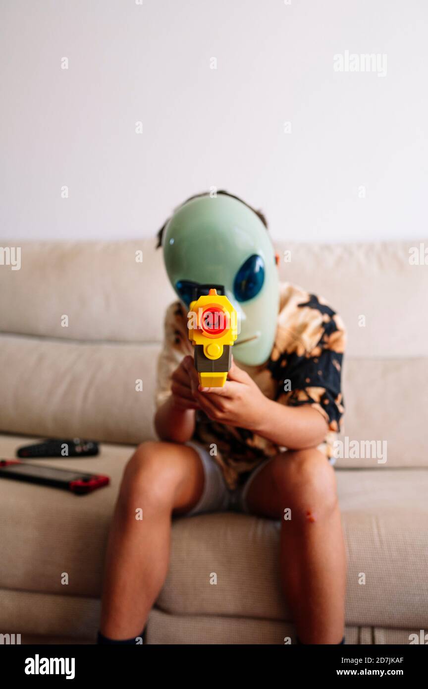 Boy wearing alien mask playing with toy gun while sitting on sofa in ...