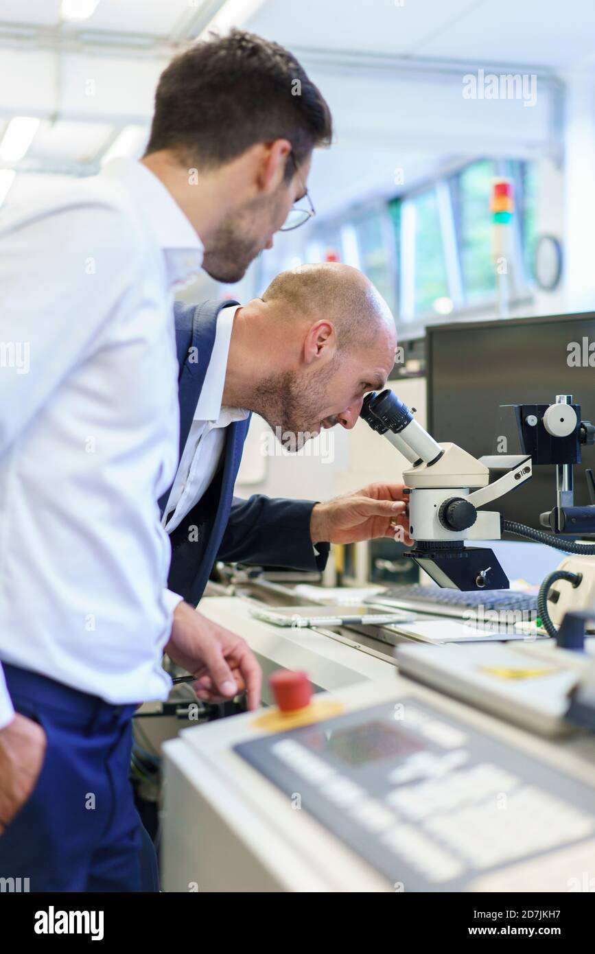 Young technician standing by male colleague looking through microscope ...
