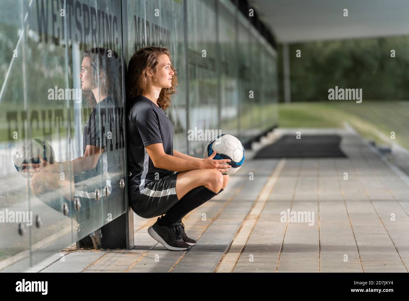 Thoughtful female soccer player holding ball while crouching by glass ...