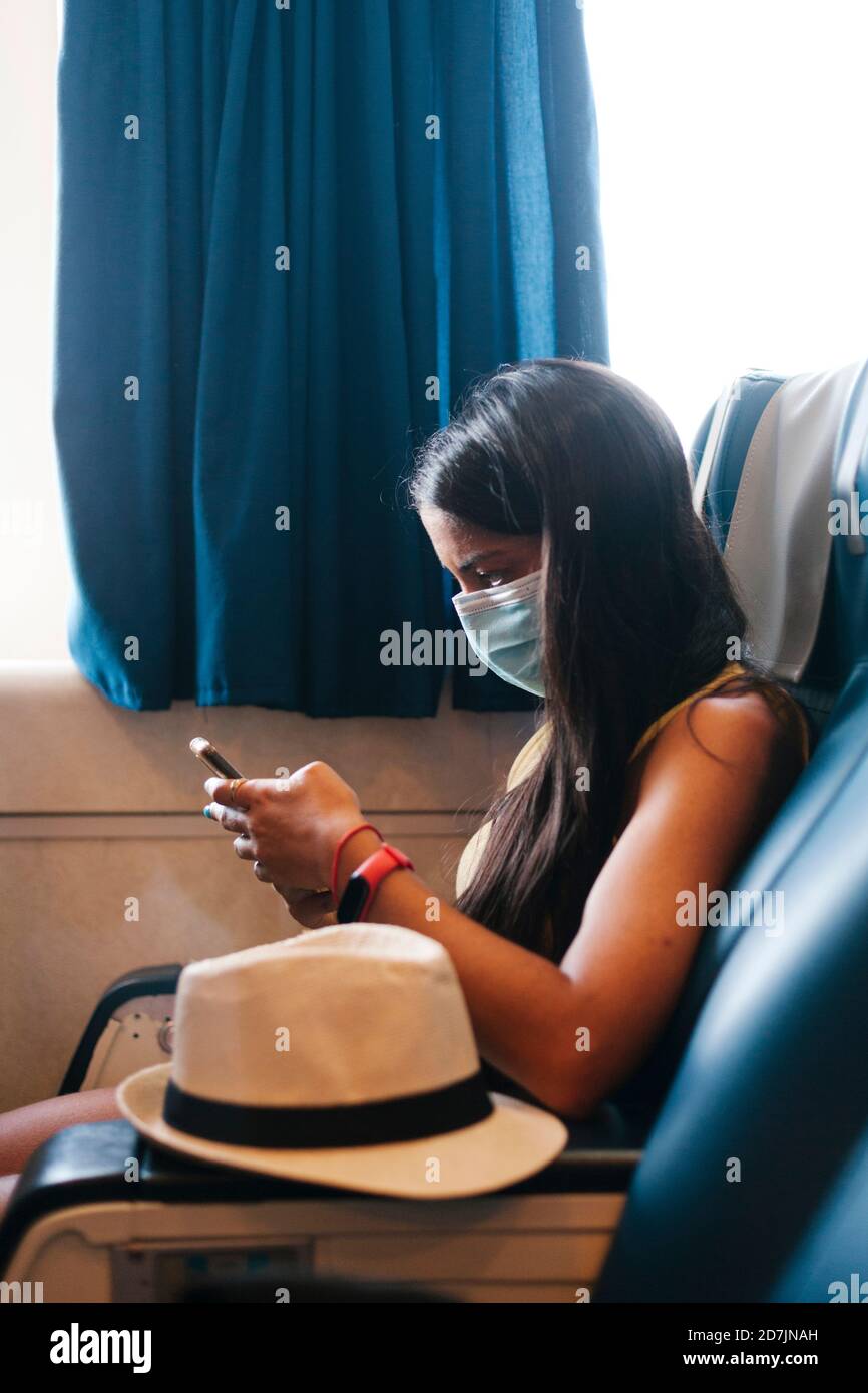 Mid adult woman wearing mask using mobile phone while sitting on seat ...