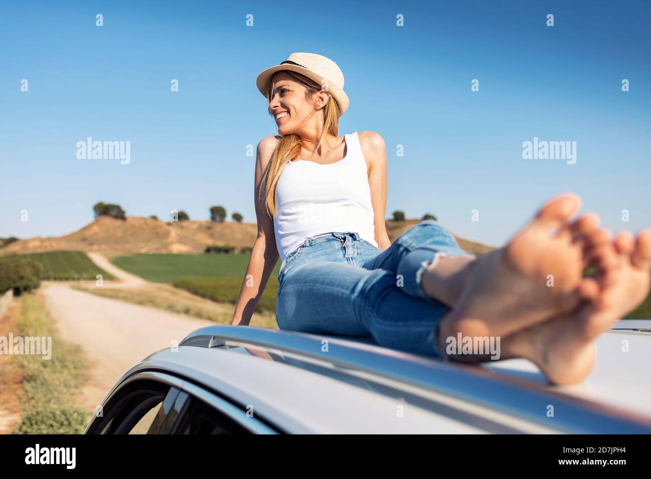 Smiling young woman with legs crossed at ankle sitting on car roof ...