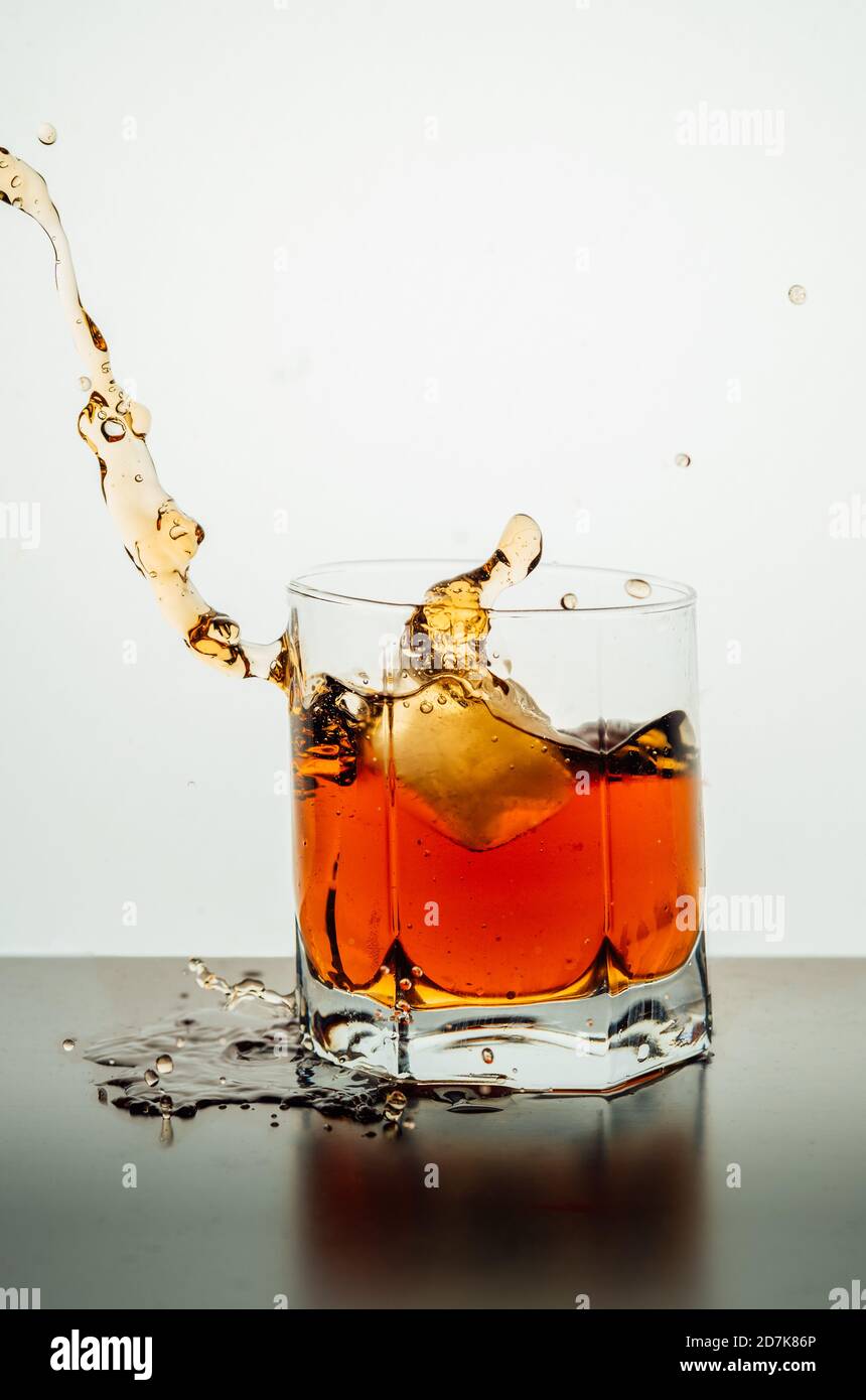 A glass of splashing whiskey with an ice piece on a reflective surface ...