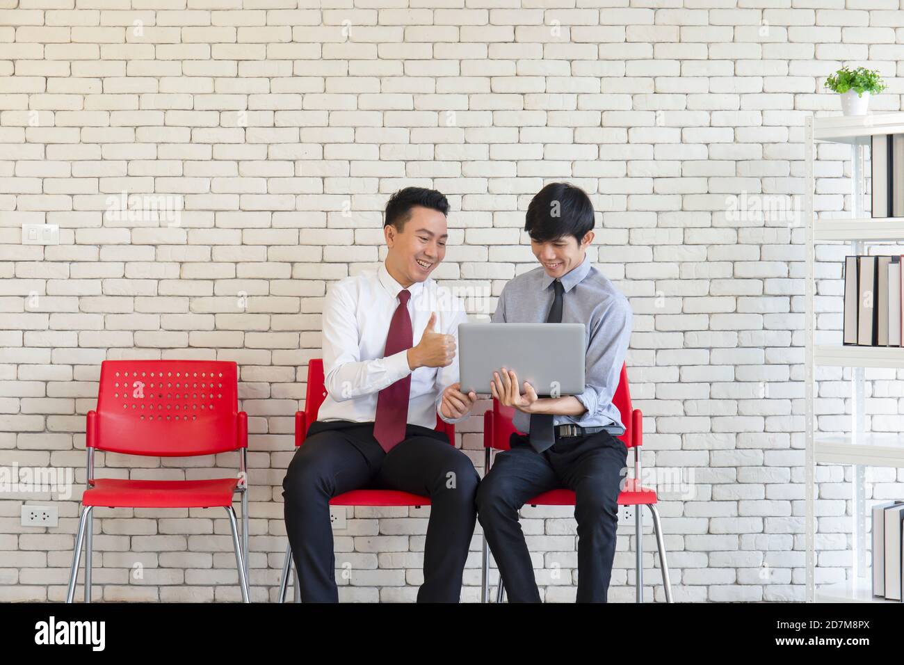Two Asian men sit on red plastic chairs waiting for a job interview ...