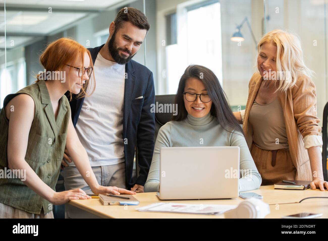 Pretty Asian businesswoman looking at laptop display among her happy ...