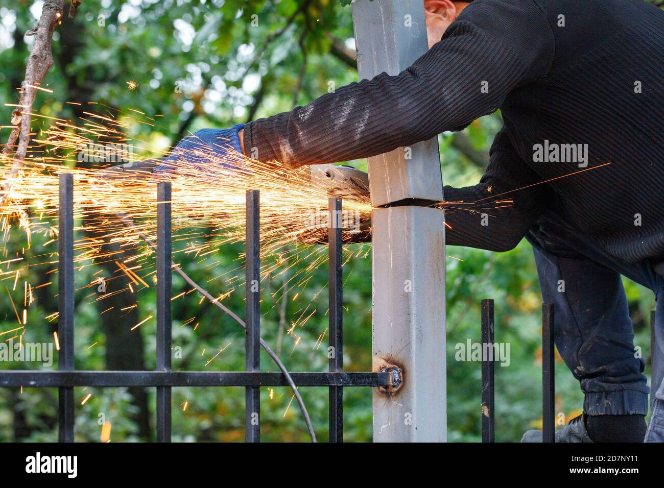 A worker cuts a metal post with a disc angle grinder, creating a bright ...
