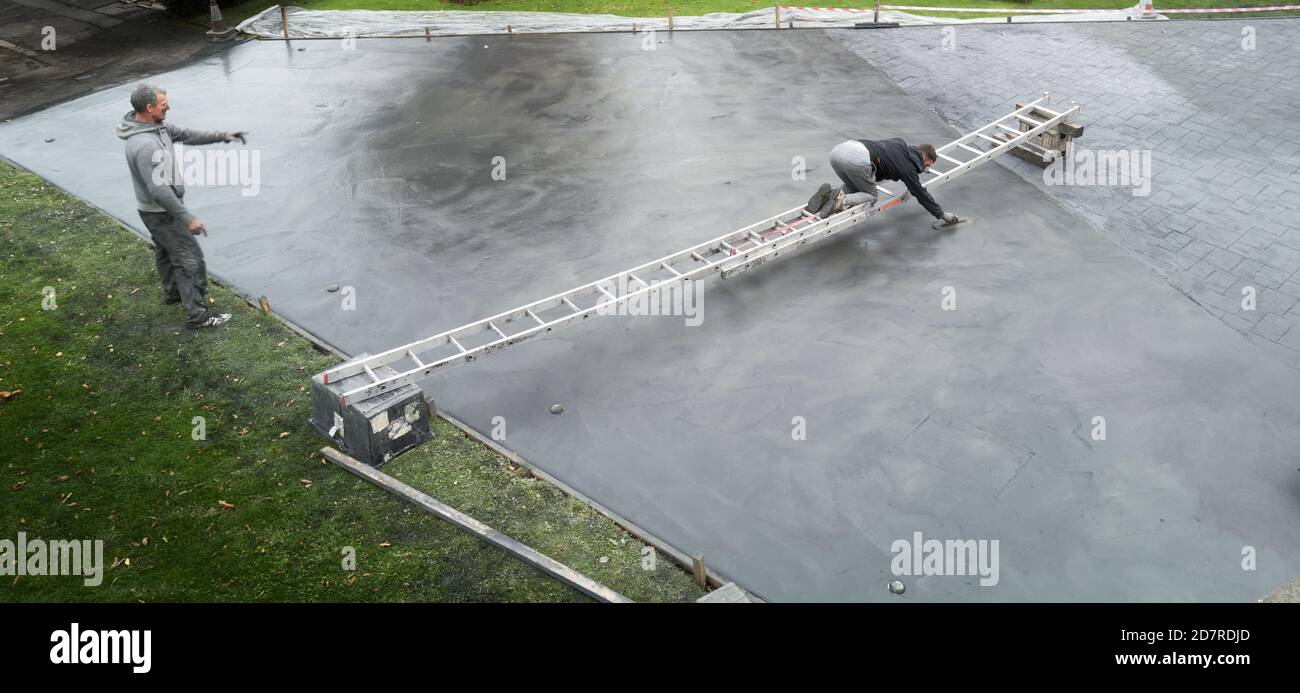 Workmen use a ladder to screed freshly laid concrete on a new driveway ...