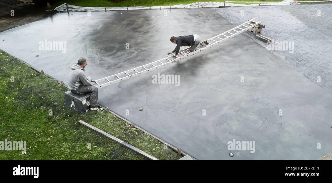 Workmen use a ladder to screed freshly laid concrete on a new driveway ...