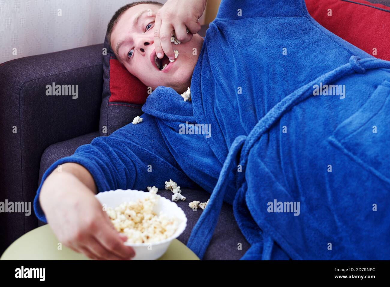 Fat man in a robe eating popcorn while lying on the couch, obesity and ...