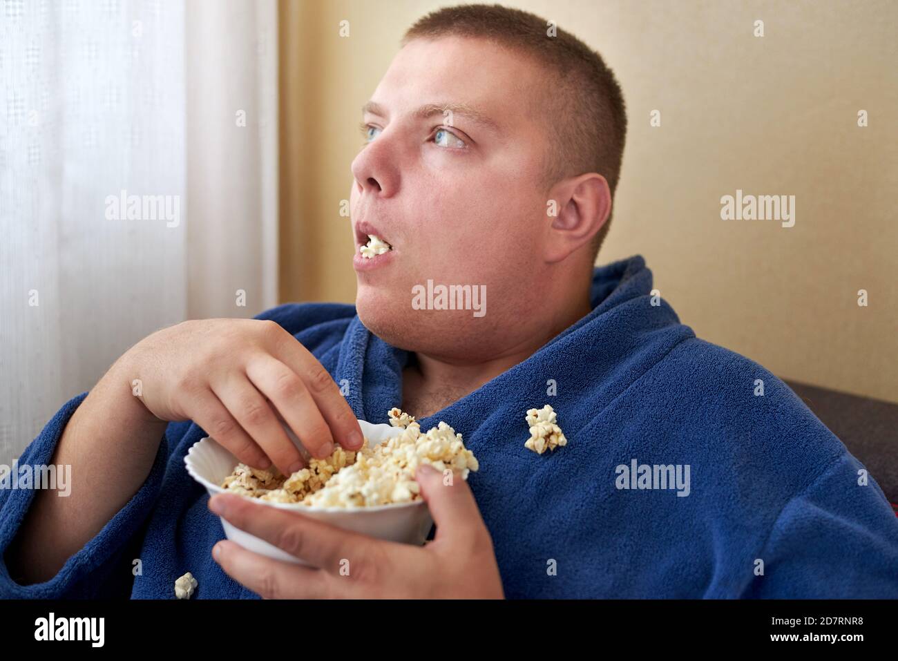 Fat man in a robe eating popcorn while sitting on the couch, obesity ...