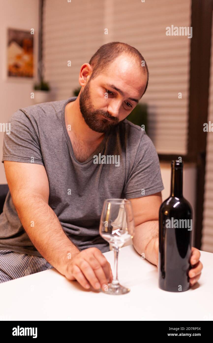 Man in dispair drinking alcohol alone sitting at table in kitchen ...