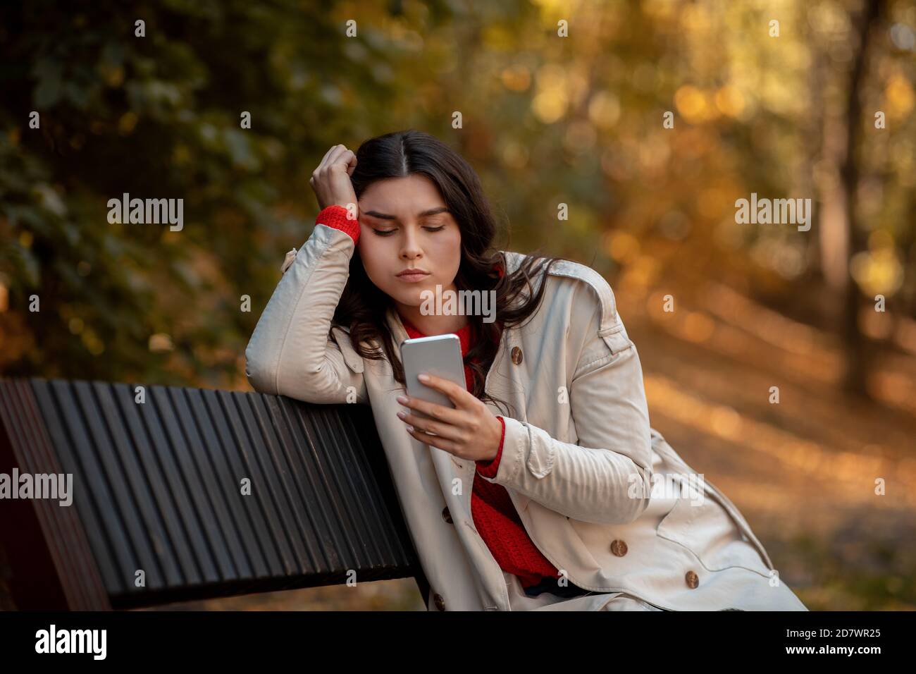 Sad young woman in autumn outfit sitting on bench at park with ...