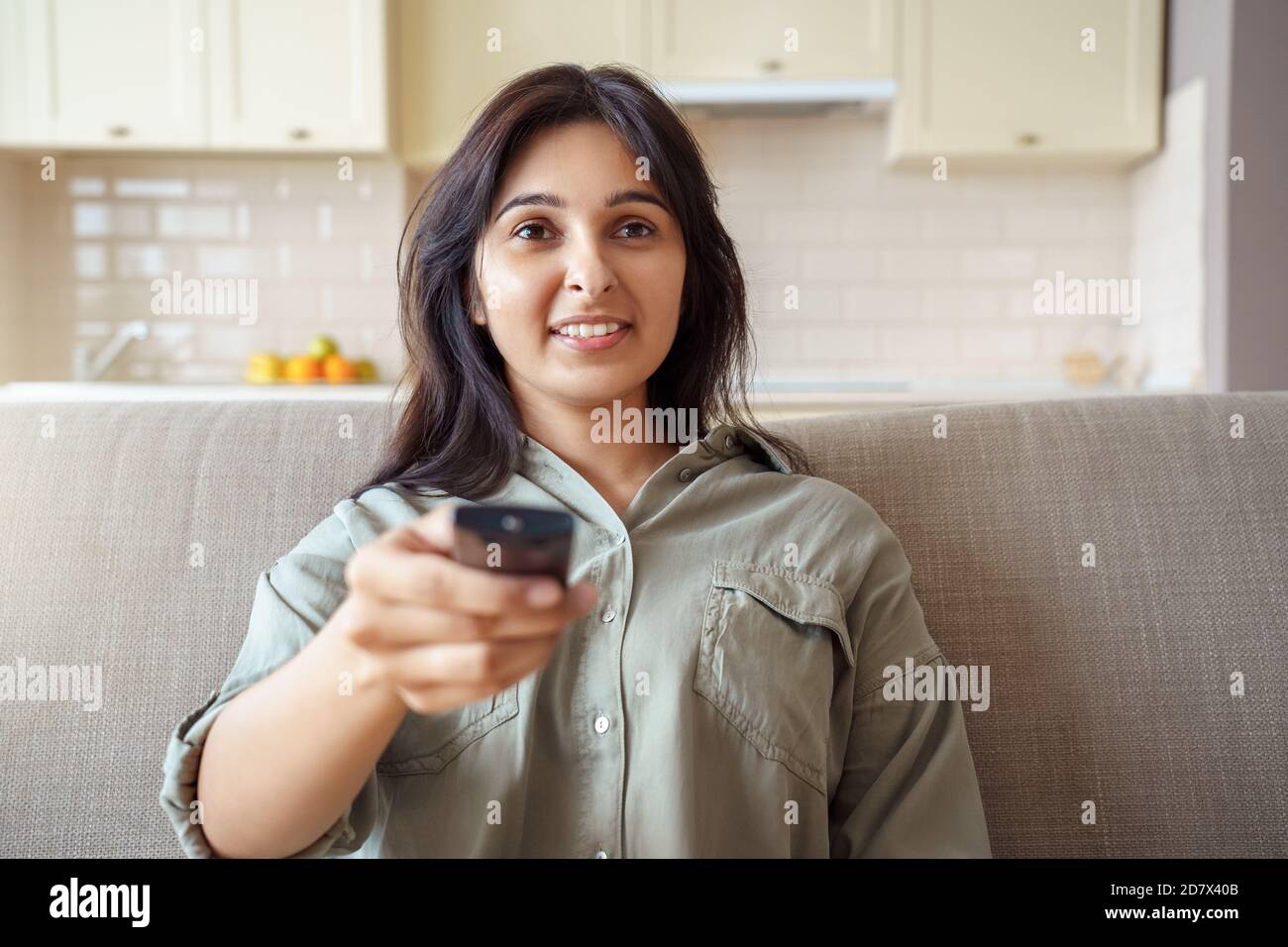 Happy indian woman sitting on sofa holding remote control watching tv ...