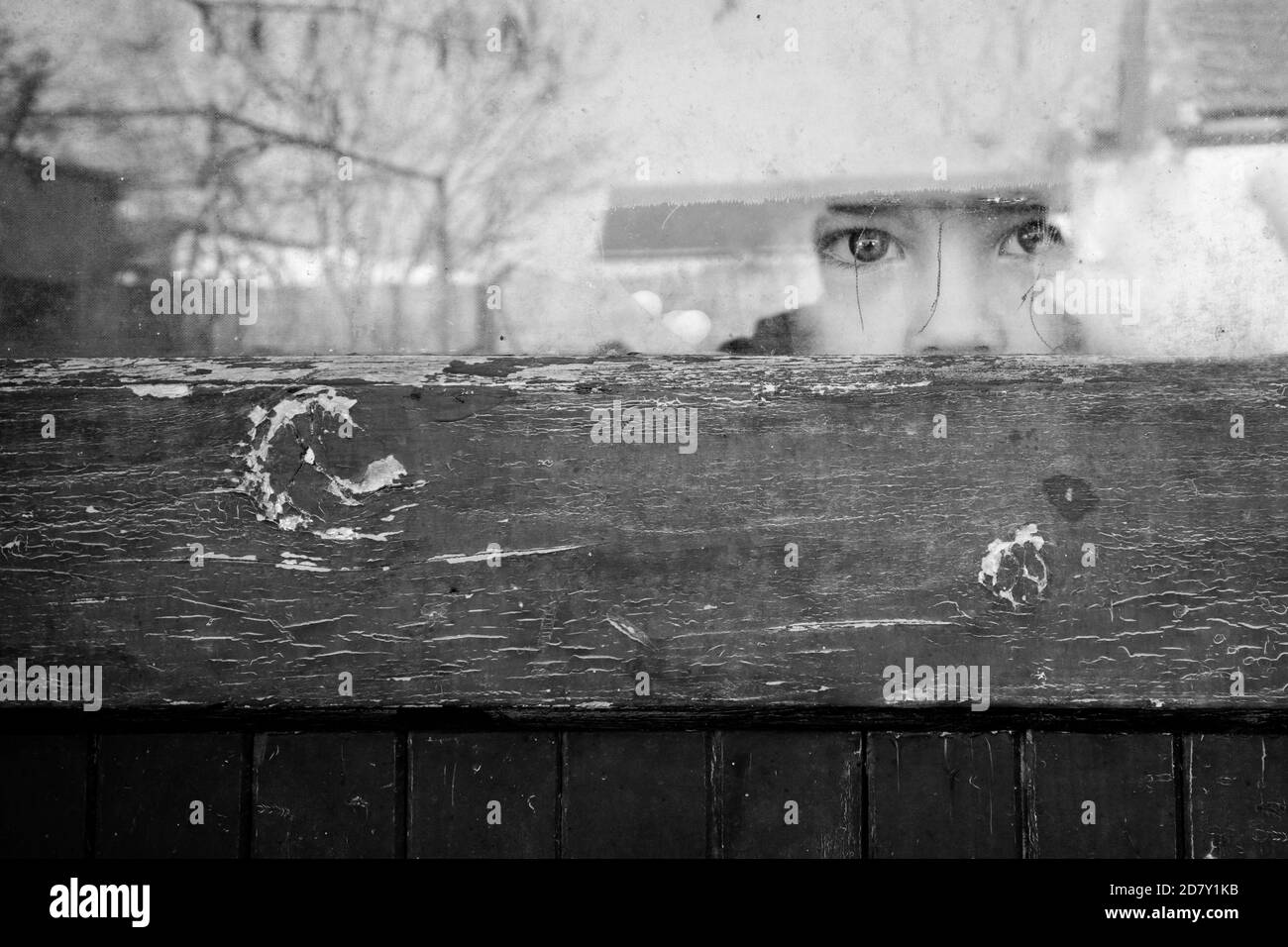 Black and white image of a child fearfully looking through a window ...