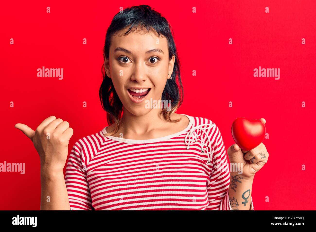 Young woman holding heart pointing thumb up to the side smiling happy ...
