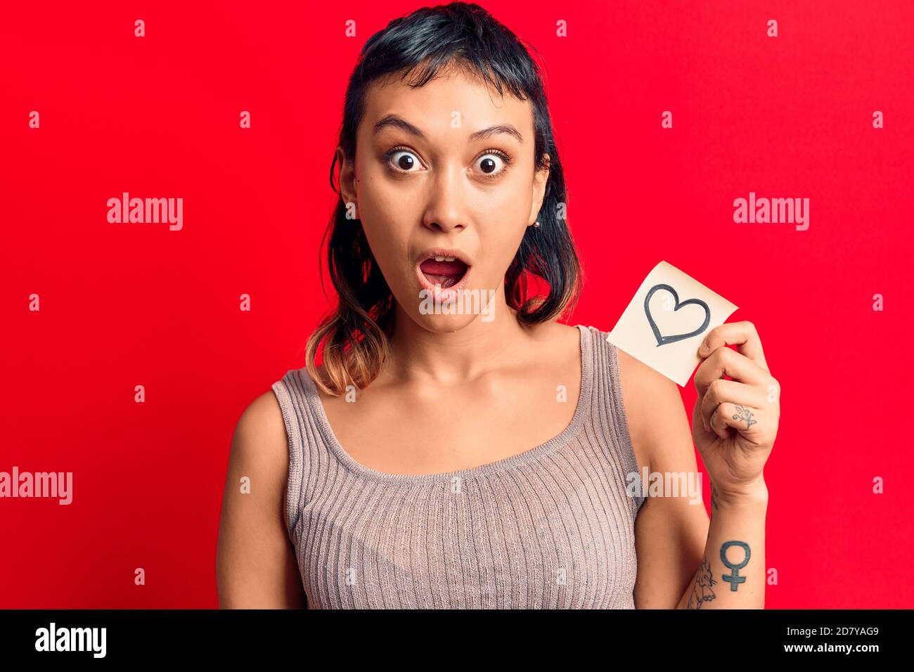 Young woman holding heart reminder scared and amazed with open mouth ...