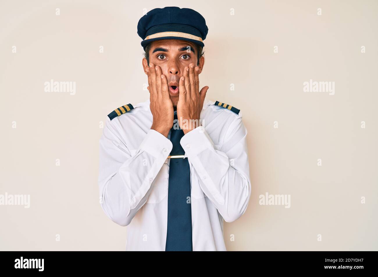 Young hispanic man wearing airplane pilot uniform afraid and shocked ...