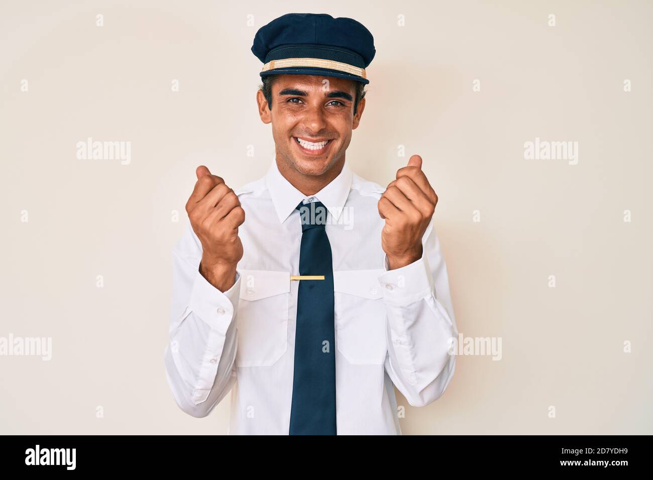 Young hispanic man wearing airplane pilot uniform doing money gesture ...