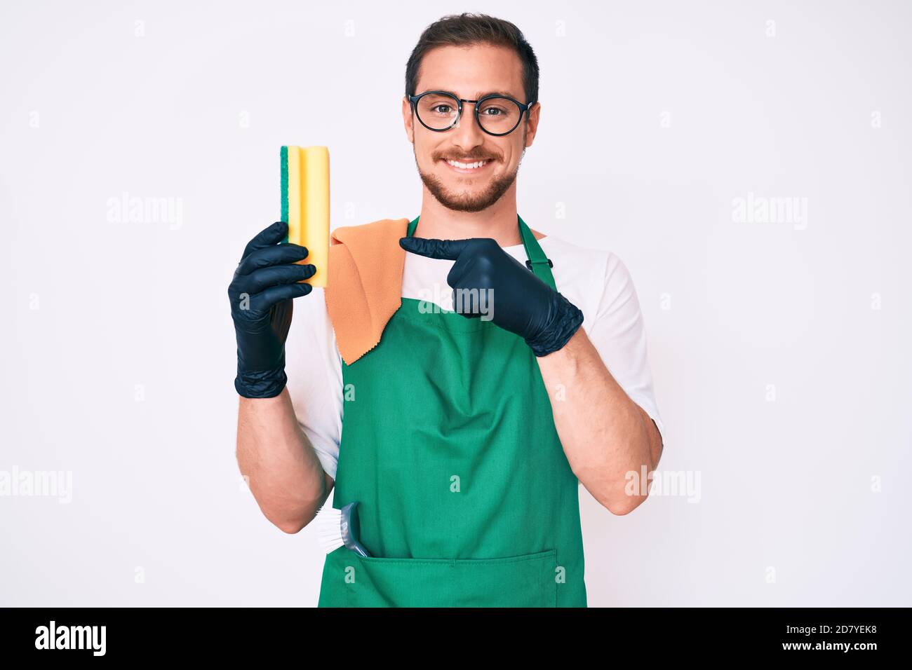 Young handsome man wearing apron holding scourer smiling happy pointing ...