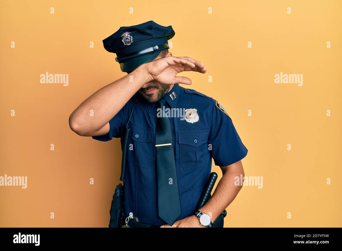 Handsome hispanic man wearing police uniform covering eyes with arm ...