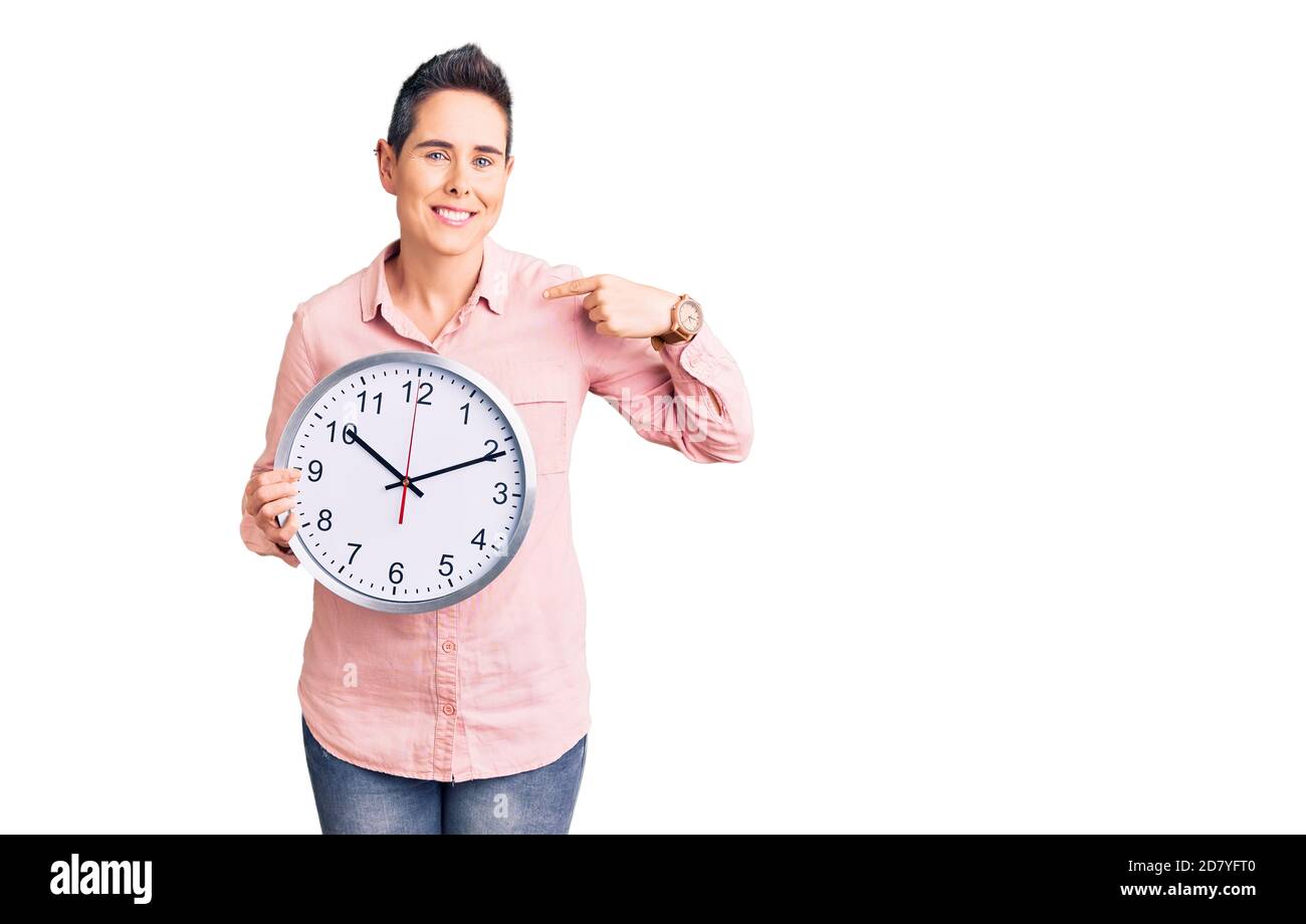 Young woman with short hair holding big clock pointing finger to one ...