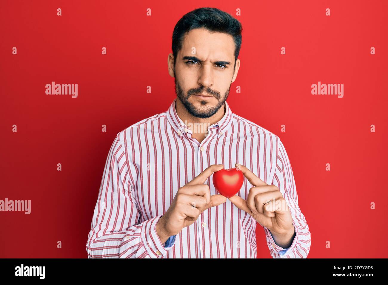 Young hispanic man holding heart skeptic and nervous, frowning upset ...