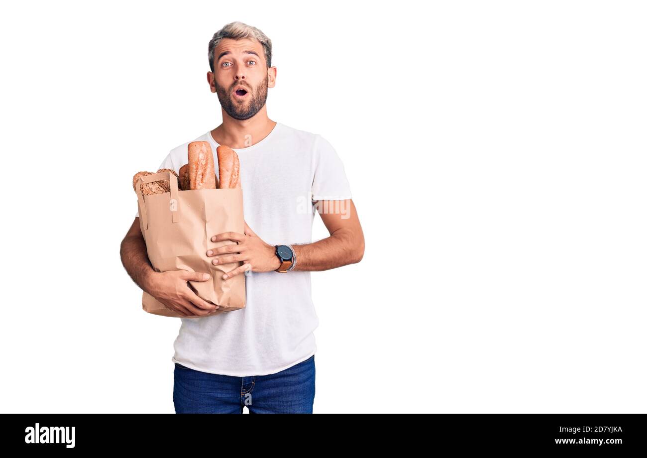 Young handsome blond man holding paper bag with bread scared and amazed ...