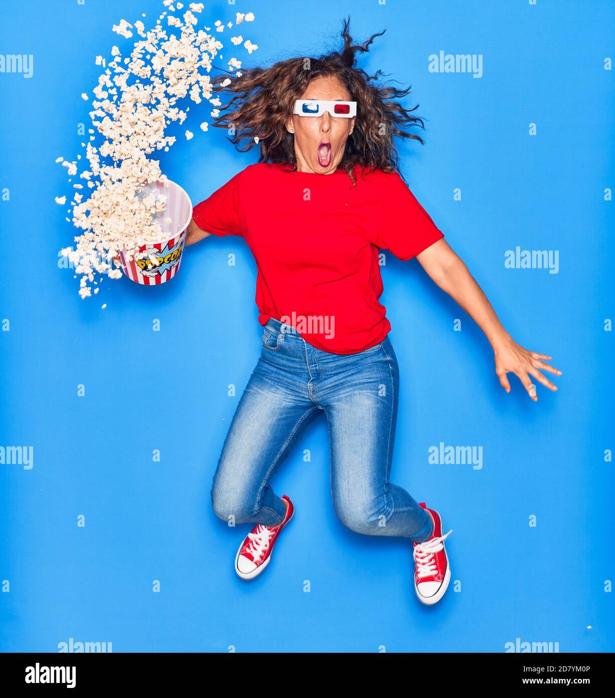 Middle age beautiful hispanic woman surprised with open mouth wearing 3d glasses. Holding bucket ...