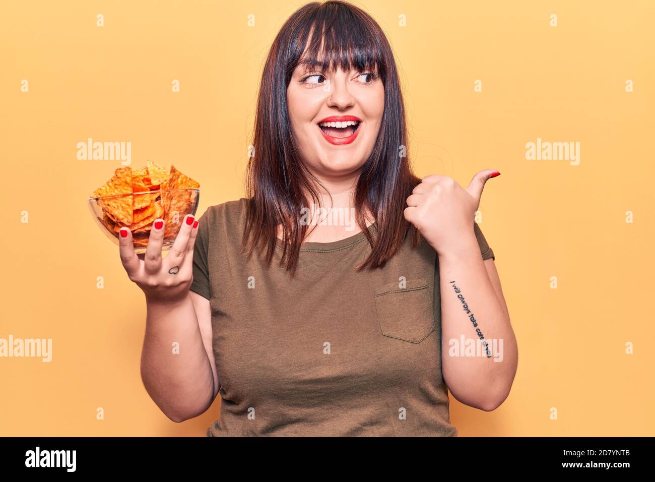 Young plus size woman holding nachos potato chips pointing thumb up to ...