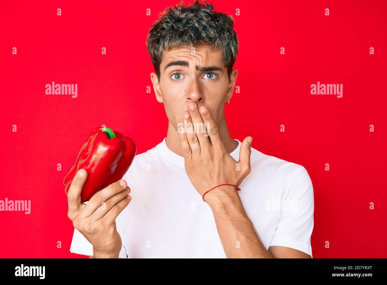 Young handsome man holding red pepper covering mouth with hand, shocked ...