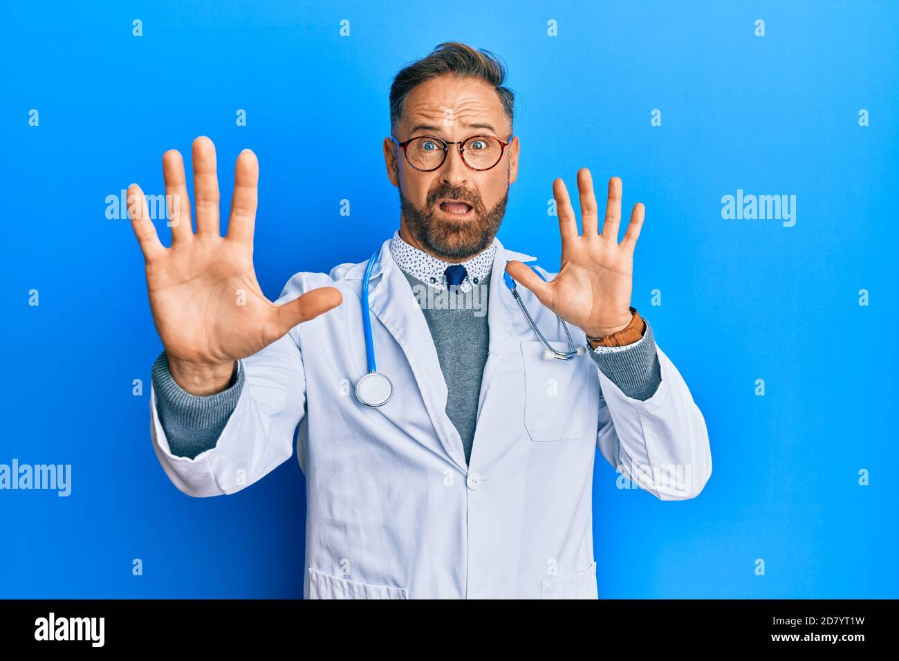 Handsome middle age man wearing doctor uniform and stethoscope afraid ...
