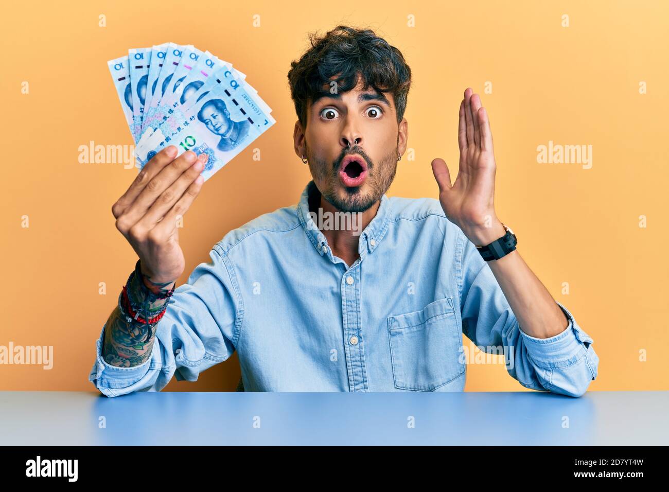 Young hispanic man holding yuan chinese banknotes sitting on the table ...