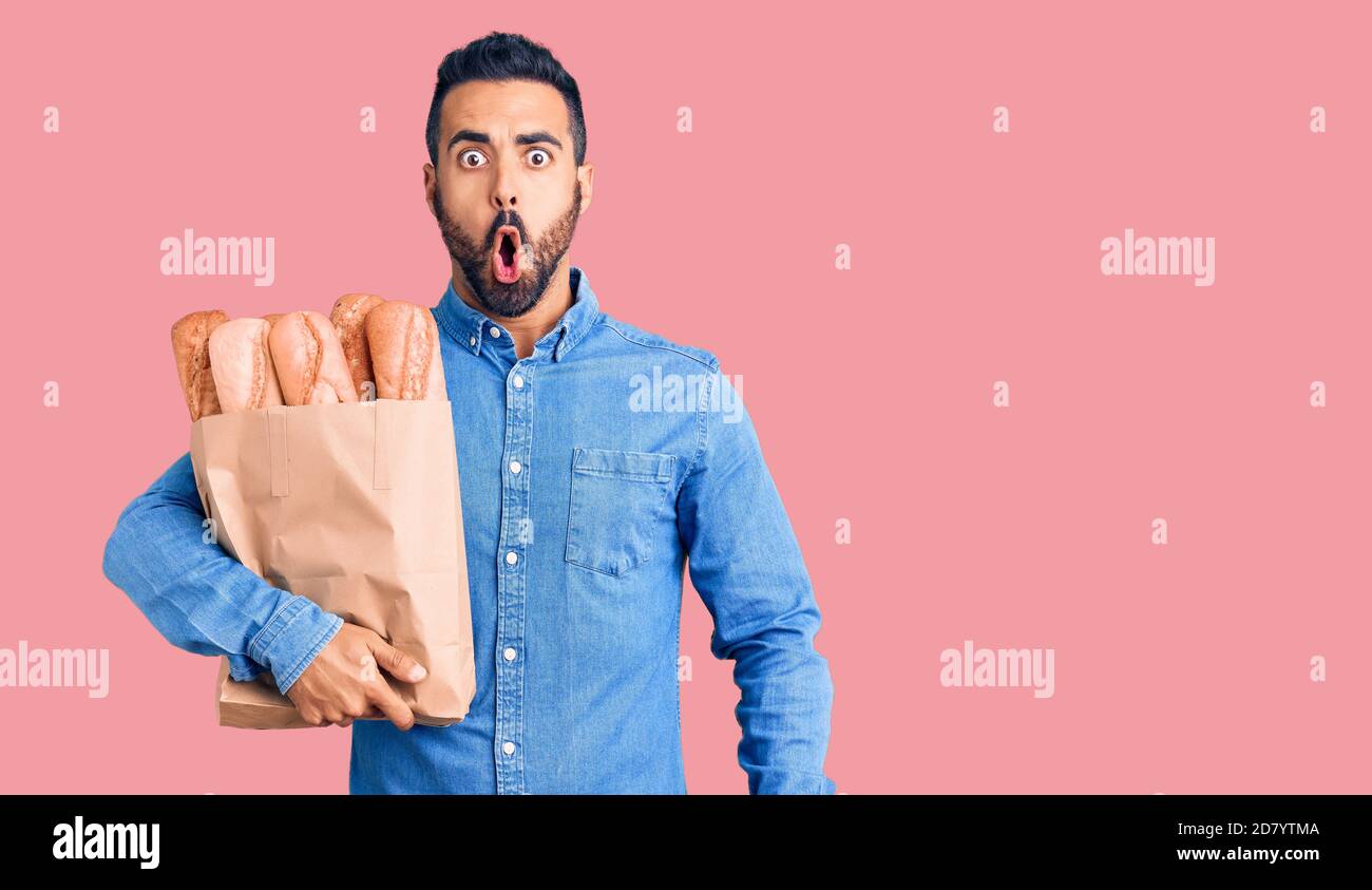 Young hispanic man holding bag with bread scared and amazed with open ...