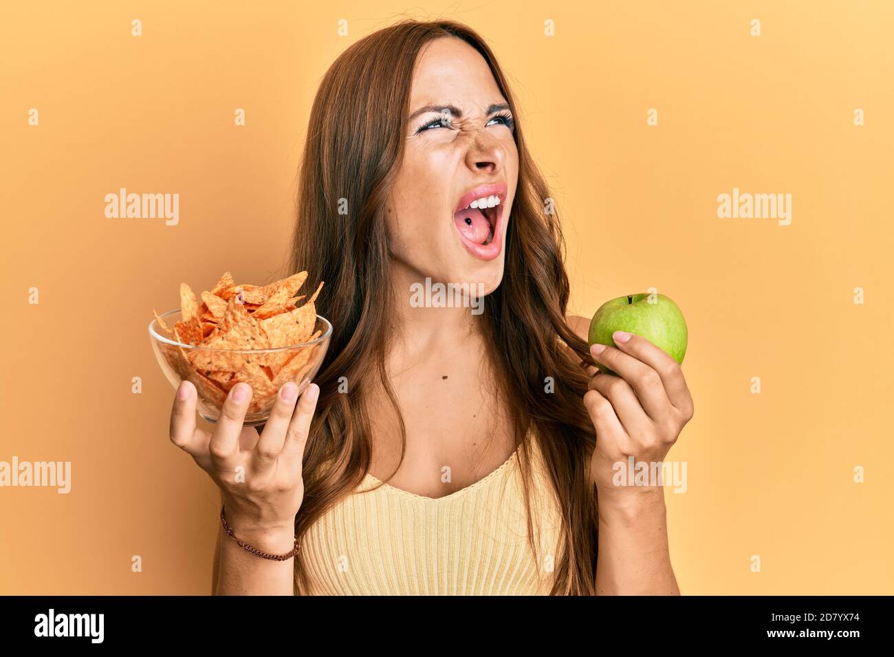 Young brunette woman holding nachos and healthy green apple angry and ...