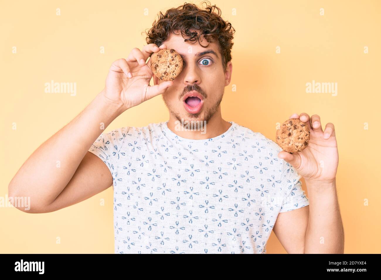 Young caucasian man with curly hair holding cookie afraid and shocked ...
