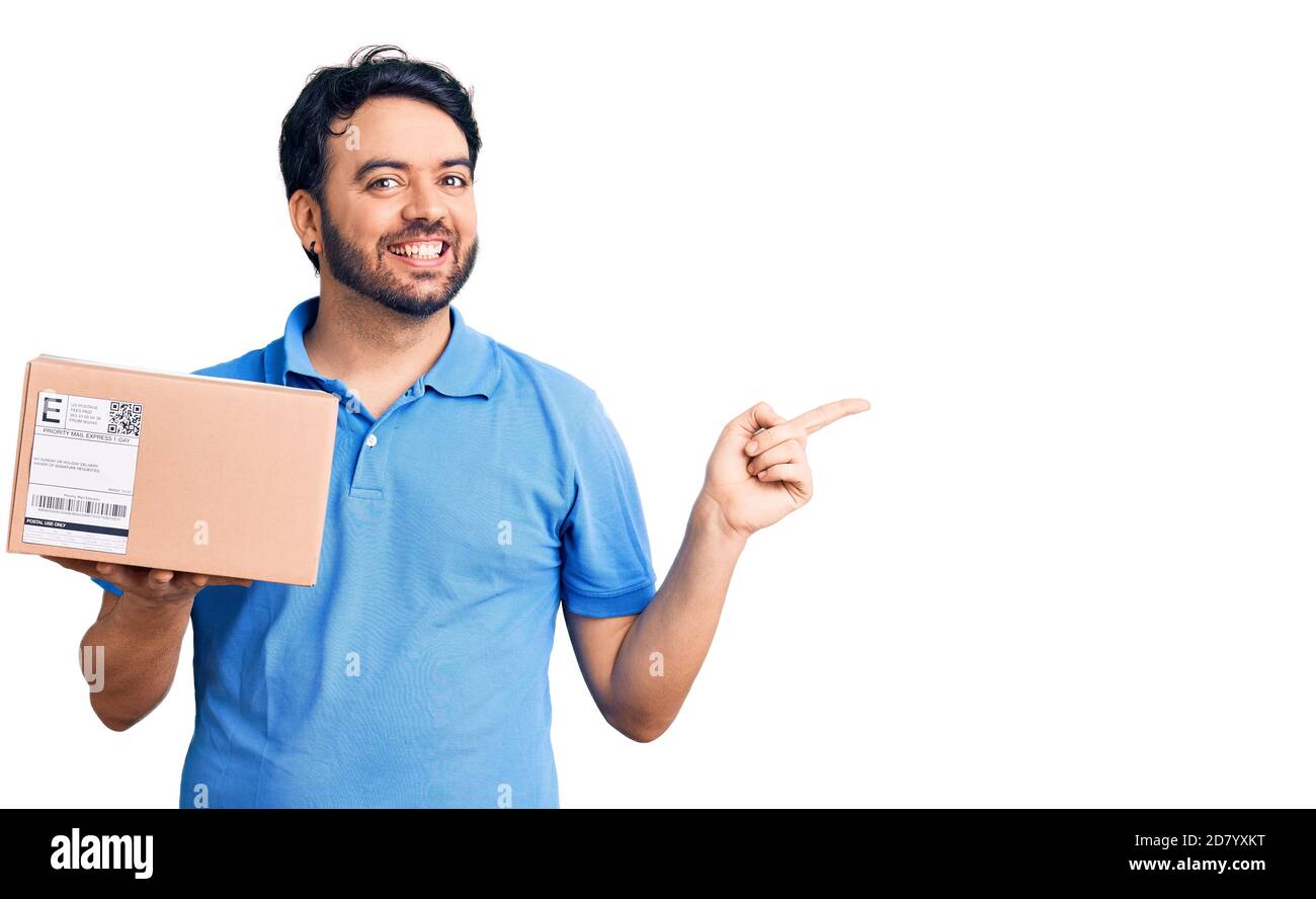 Young hispanic man holding delivery package smiling happy pointing with ...