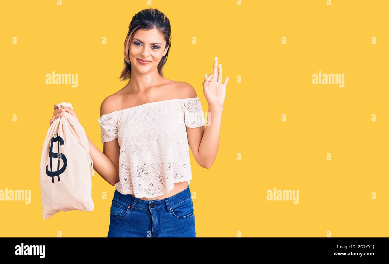 Young beautiful woman holding money bag with dollar symbol doing ok ...