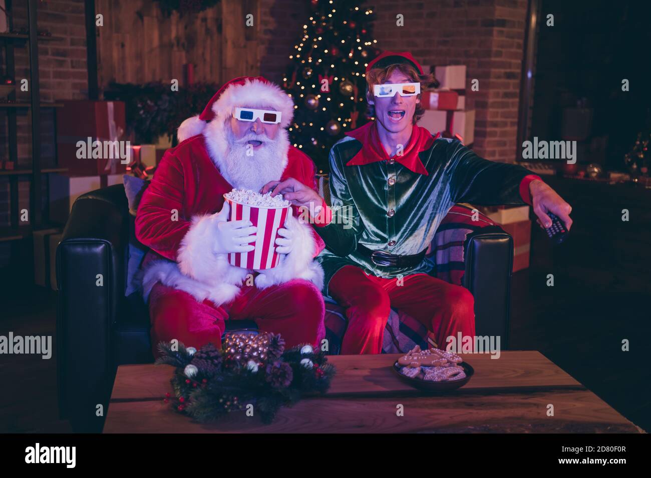 Photo portrait of santa claus and elf eating popcorn using remote ...