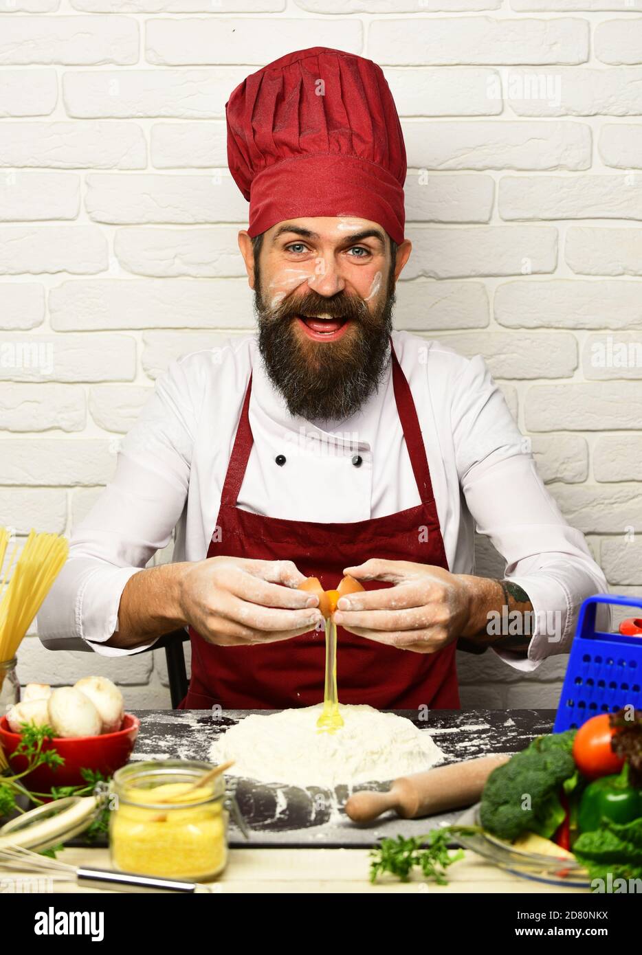 Cook with cheerful face in burgundy uniform sit by table with ...