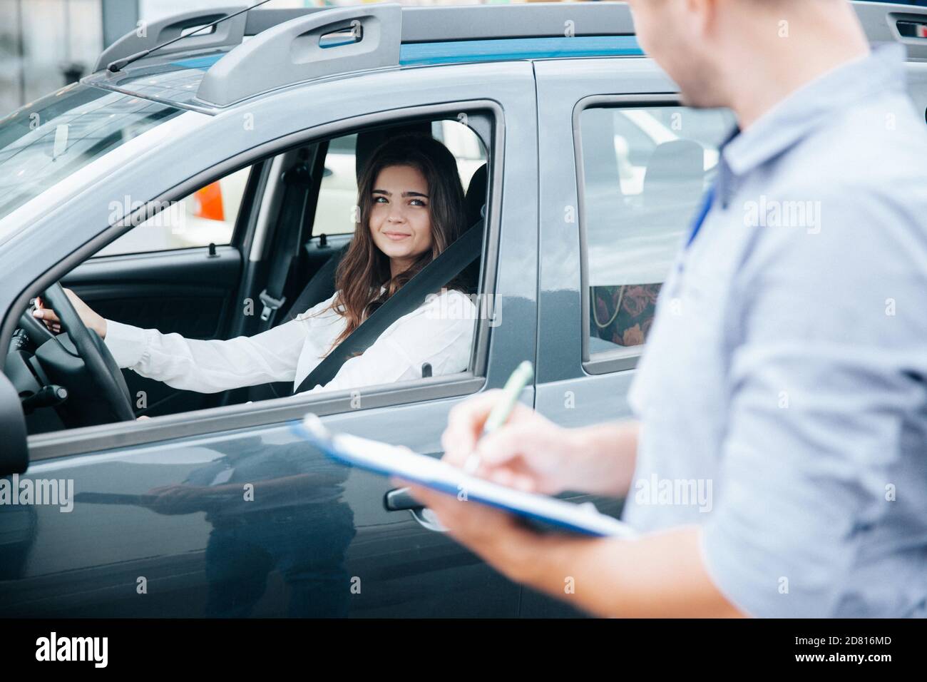 Pretty young woman sits in gray car holding a wheel and smiles to male ...