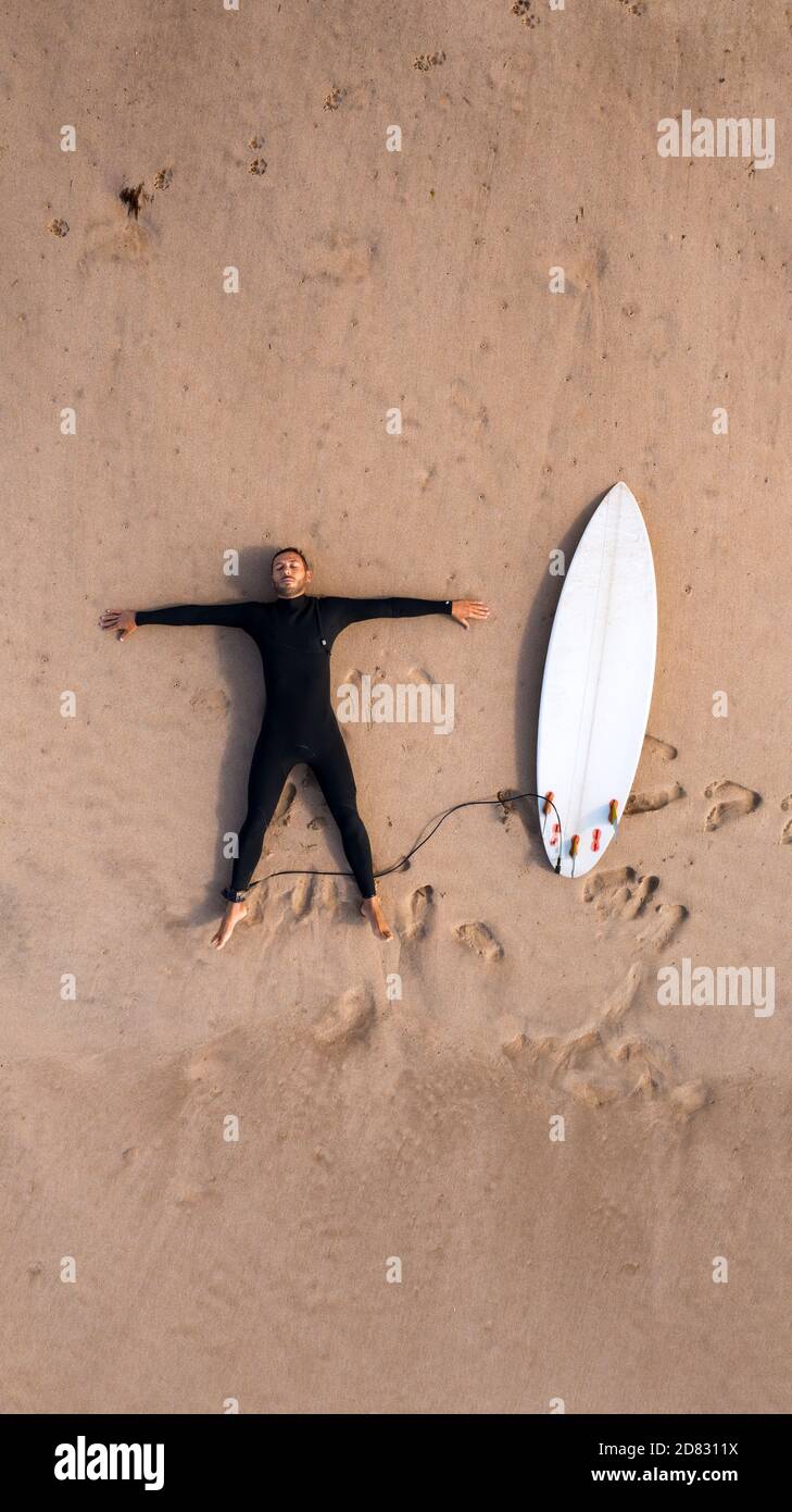 Top view of a man surfer in wetsuit with a surfboard lying on the sand ...