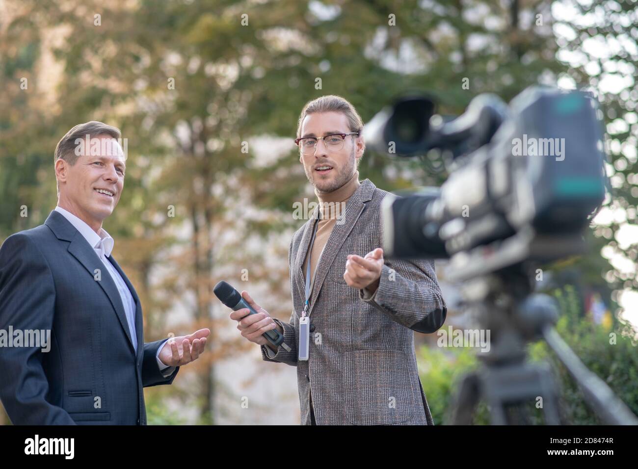 Fair-haired male journalist interviewing smiling mature male in front ...