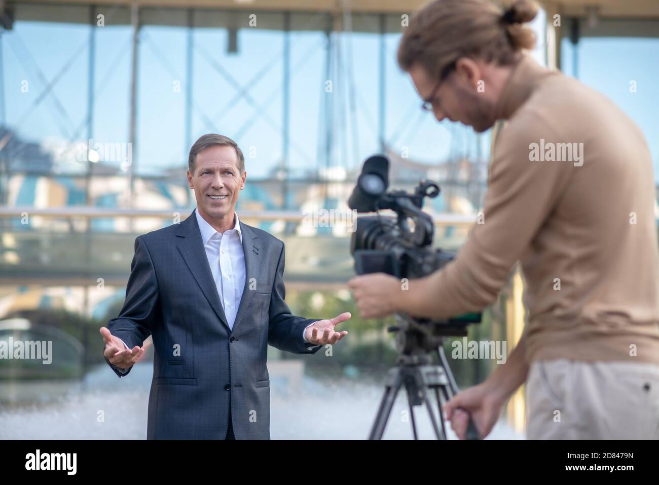 Long-haired operator filming smiling mature male reporter in front of ...