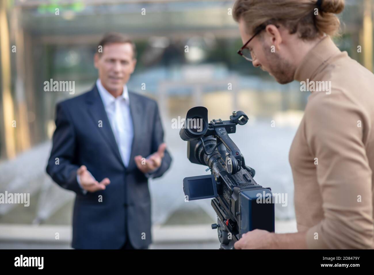Long-haired operator filming mature male reporter in front of fountain ...