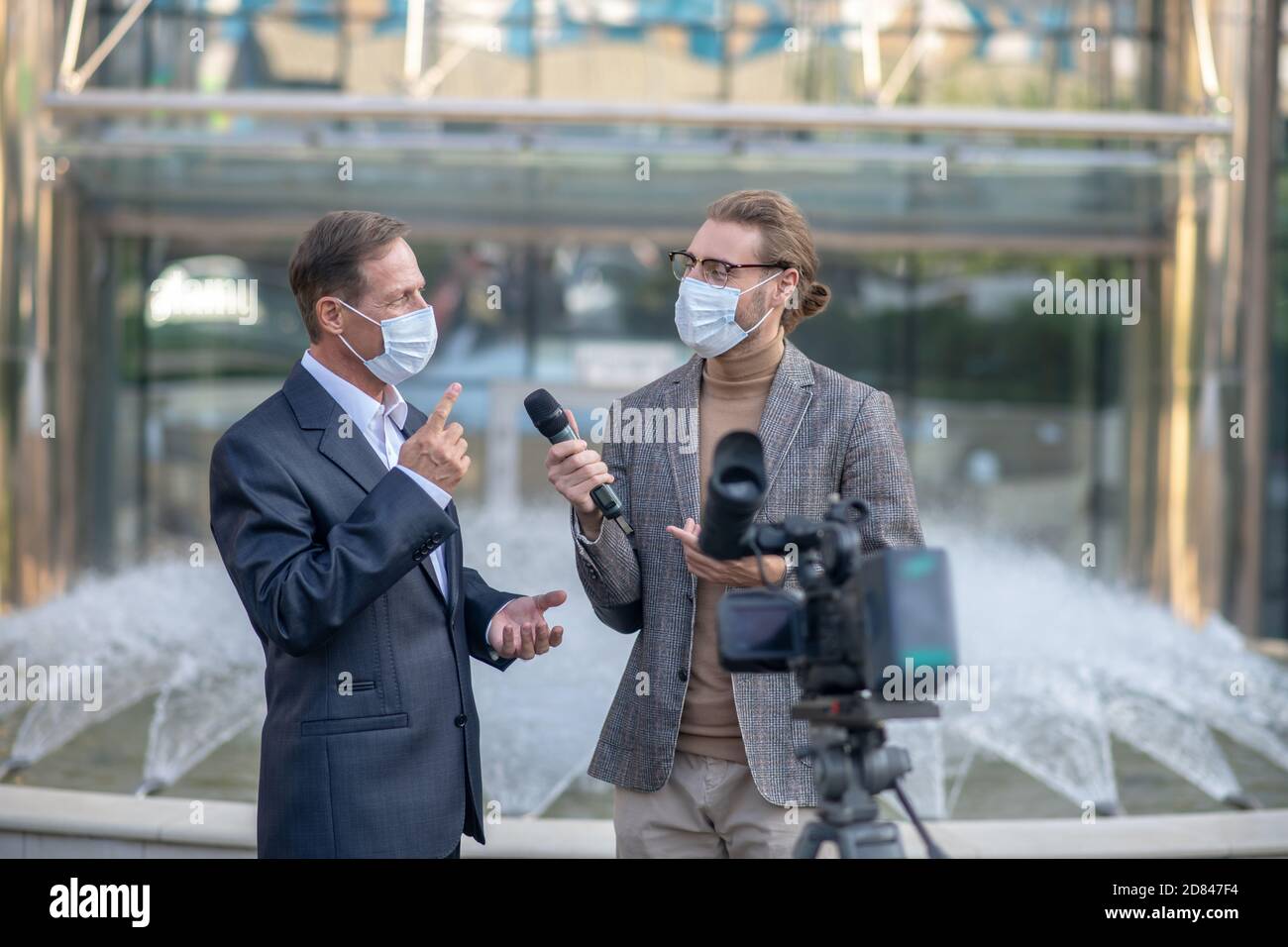 Fair-haired male journalist interviewing mature male in front of camera ...