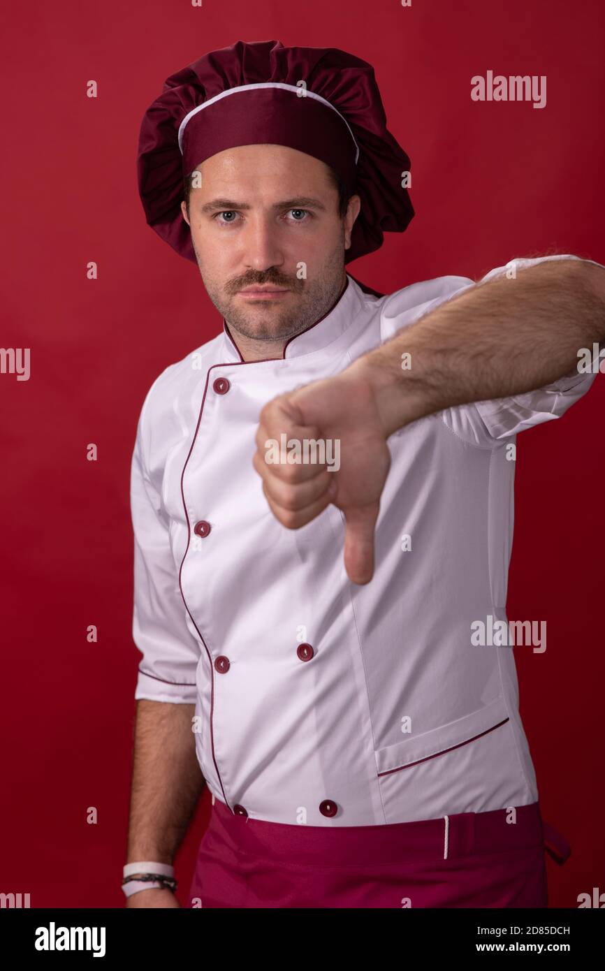 Unshaven handsome male chef shows thumb down looking at camera on red ...
