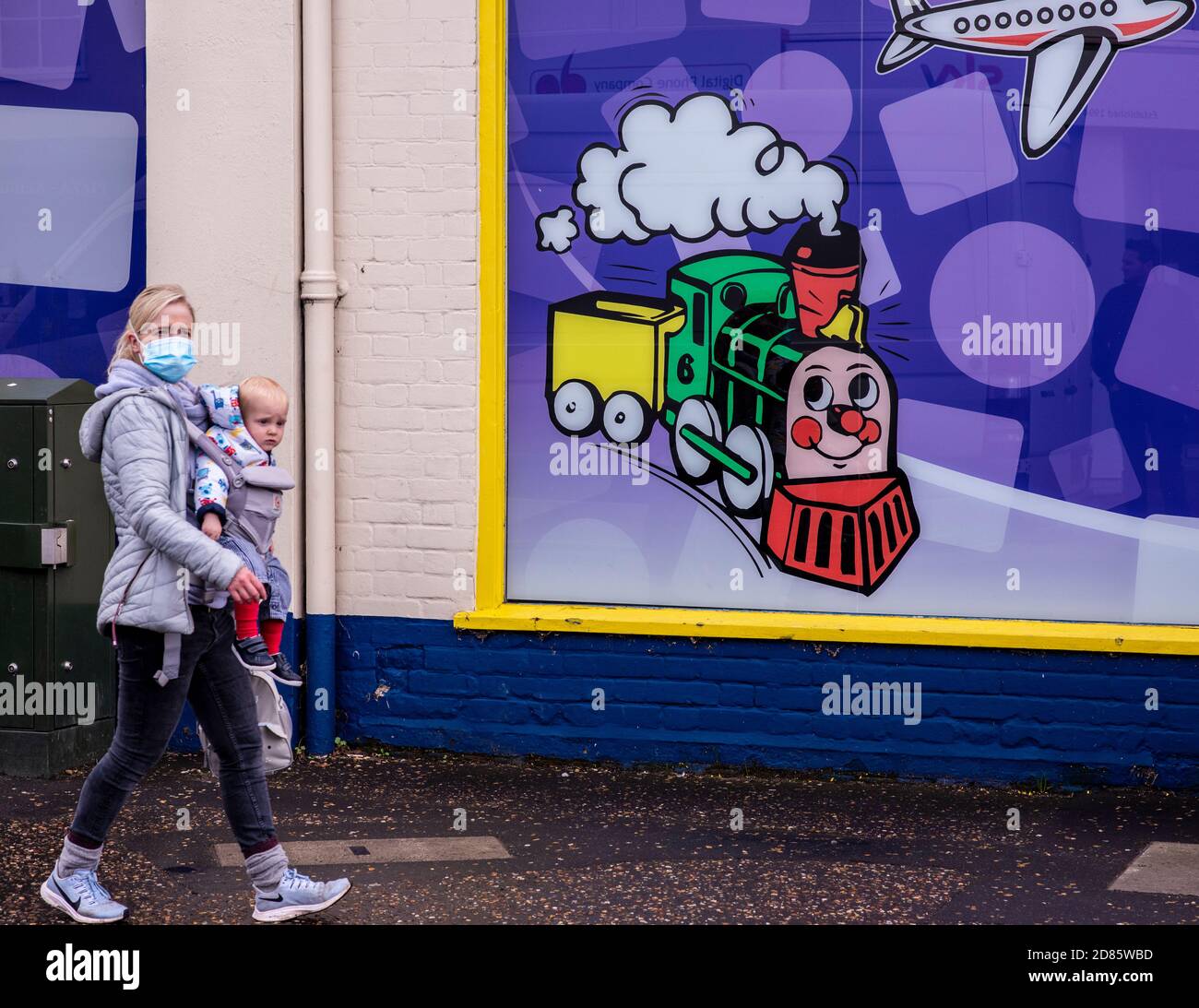 Woman wearing face mask with baby in carrier walking past toy shop with ...