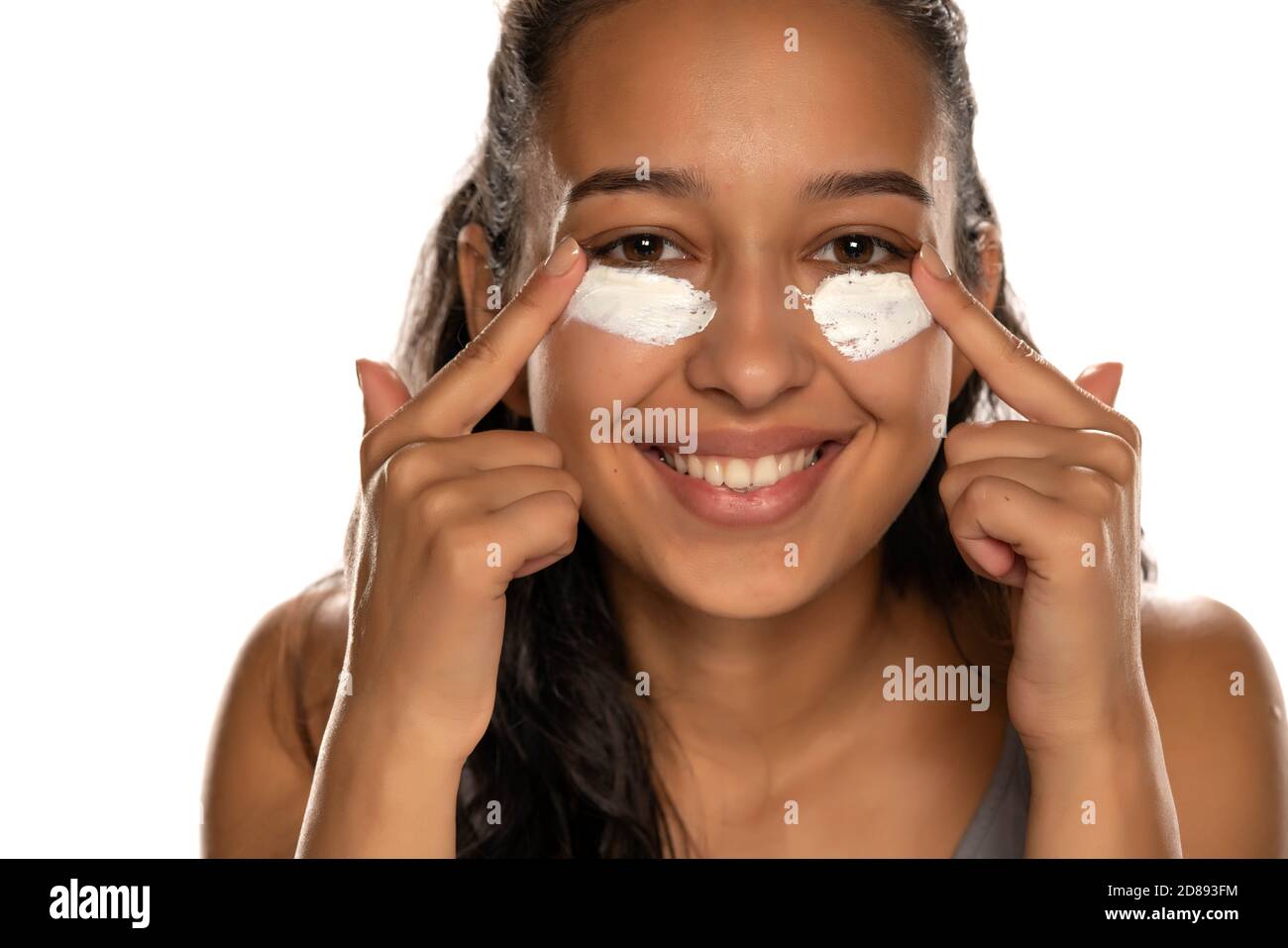 Young dark skinned woman applying a creme on her low eyelids on white ...