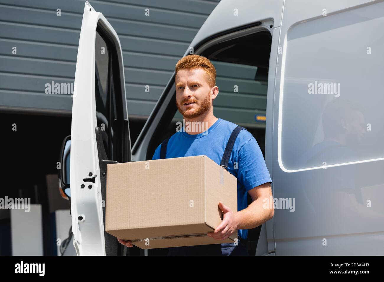 Loader in uniform carrying cardboard box near truck on urban street ...