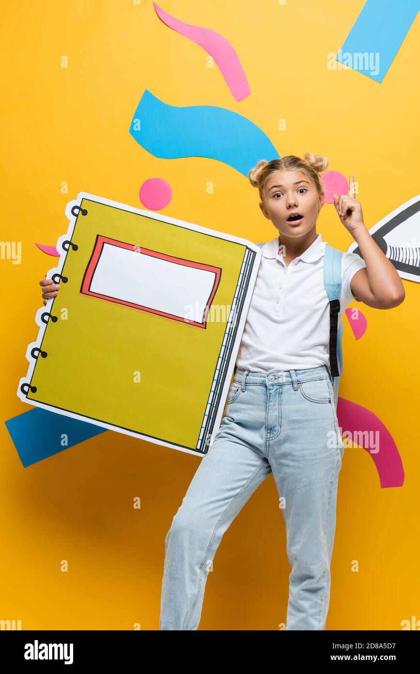 amazed schoolgirl with notebook maquette showing idea gesture on yellow ...